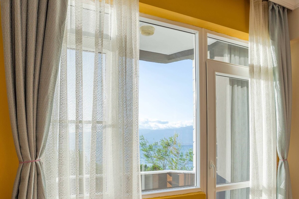 Quarto com cama, janela com cortinas e vista para montanhas.