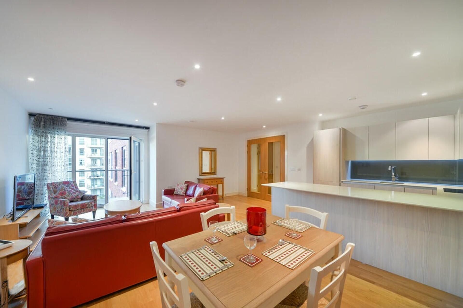 Uma sala de estar moderna com um sofá vermelho, uma mesa de jantar posta para quatro pessoas e uma área de cozinha com uma grande janela.