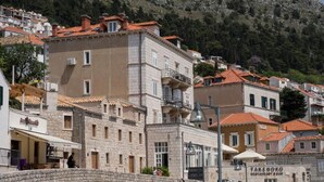 Uma vila costeira com edifícios de pedra, um restaurante chamado 'TAKENOKO' e montanhas ao fundo.