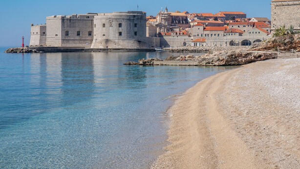 Uma vila costeira com edifícios históricos de pedra, mar azul cristalino e uma praia arenosa.