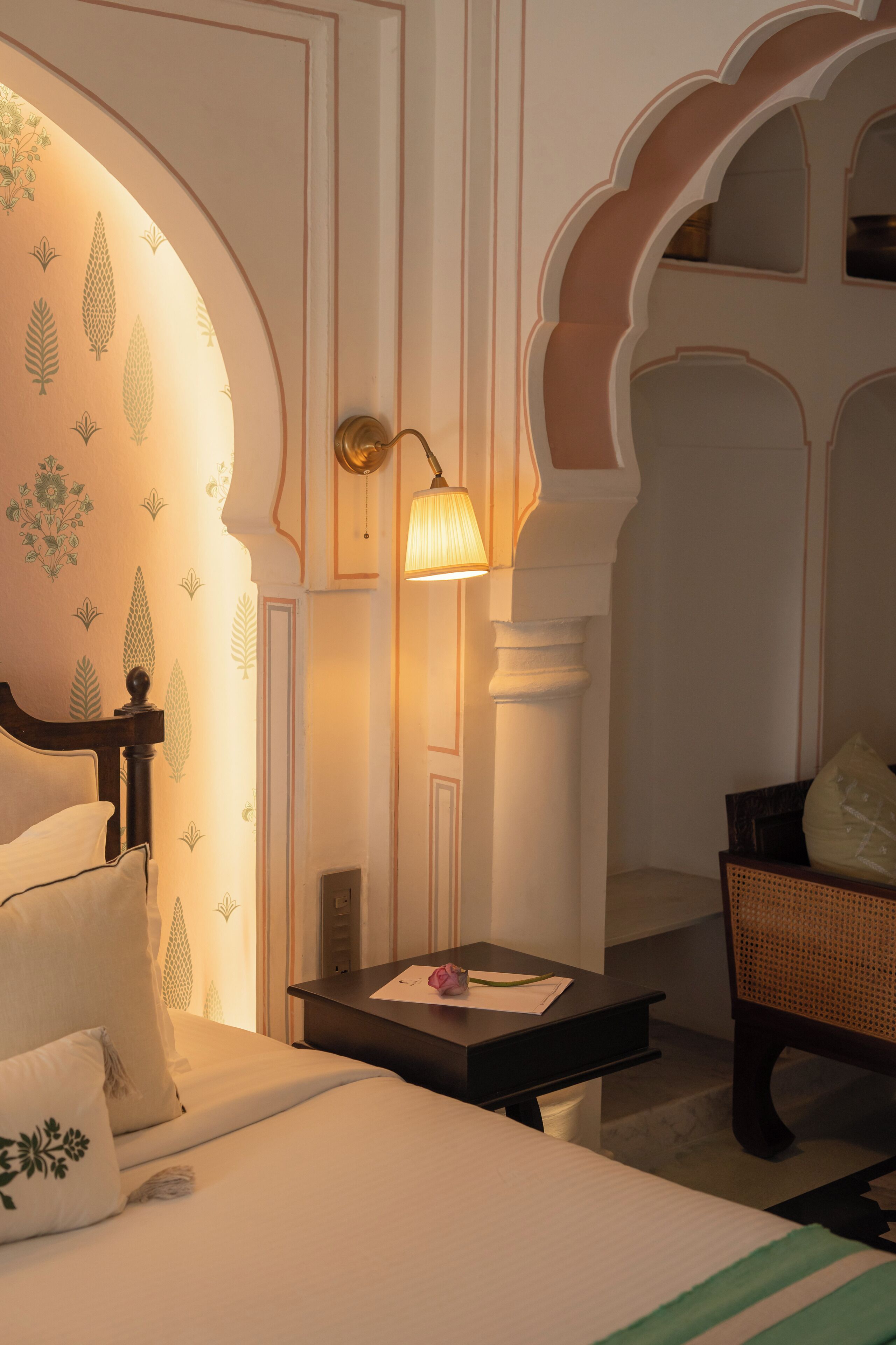 Quarto de hotel com cama, mesa de cabeceira, luminária, cadeira e padrões decorativos nas paredes.