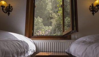 Quarto com duas camas, janela com vista para árvores e flores, e arandelas na parede.