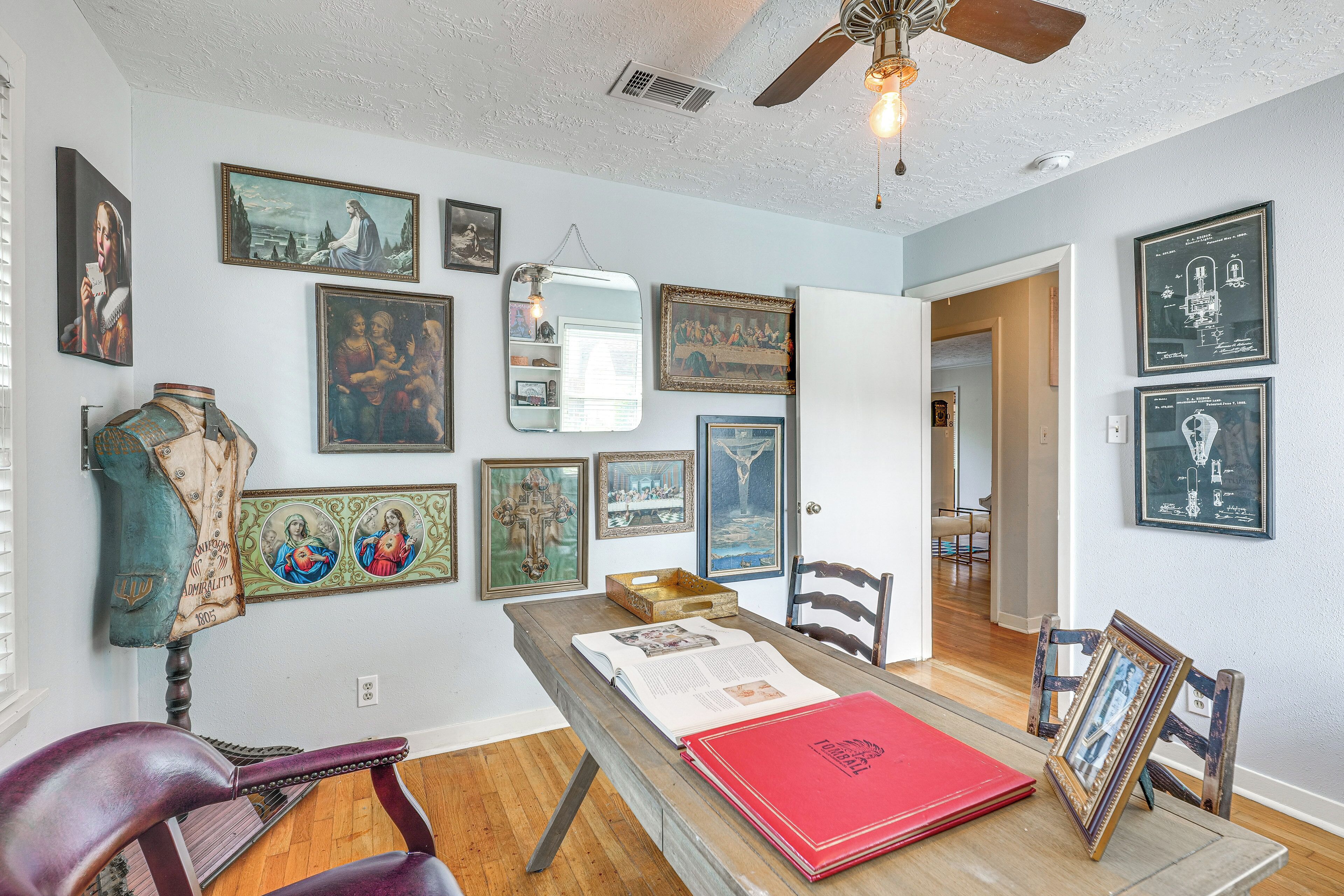 Um quarto com uma mesa de madeira, um livro vermelho e várias molduras nas paredes.