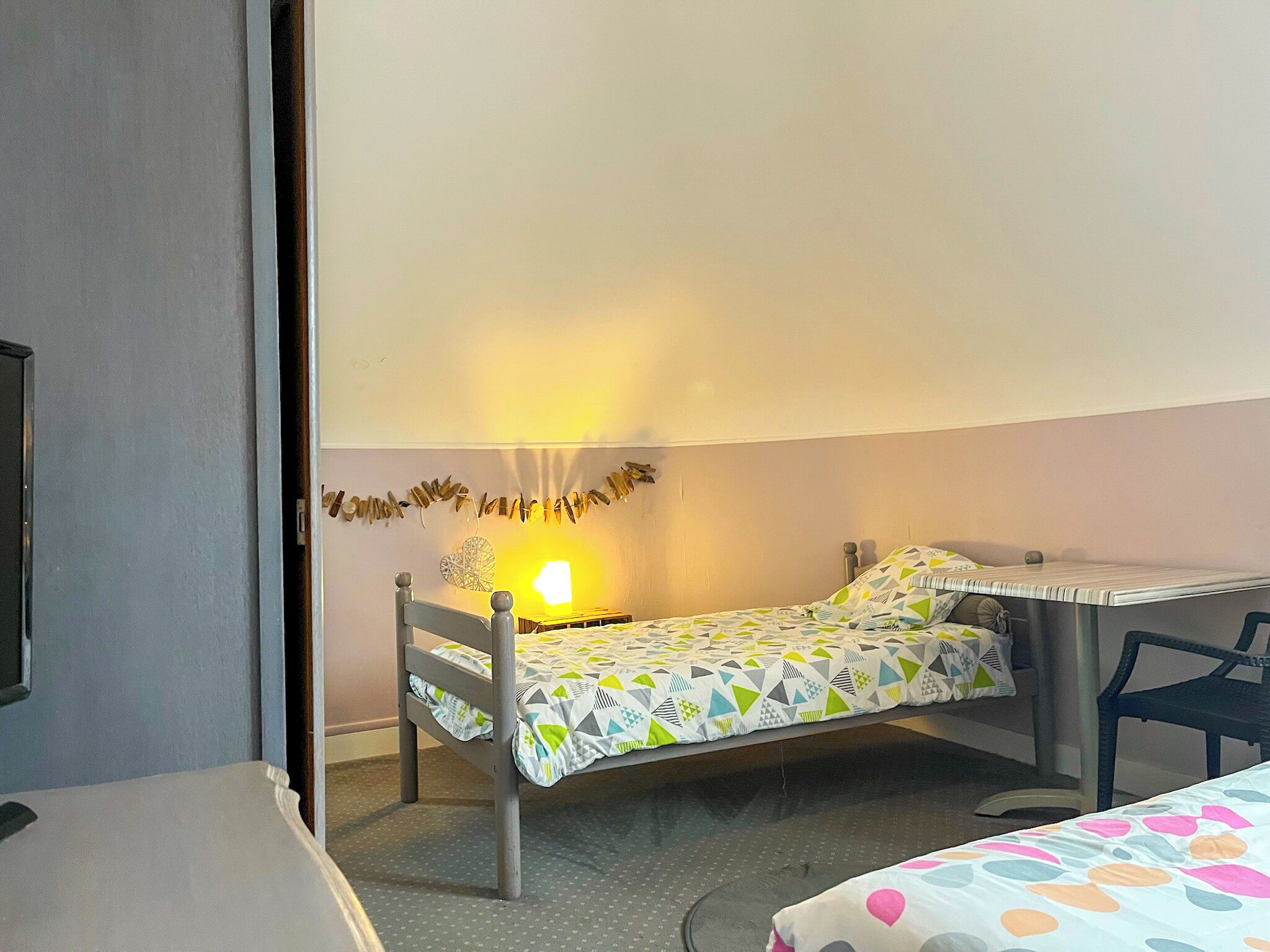 Uma cama de solteiro com colcha estampada em padrão geométrico, uma mesa de cabeceira e uma luminária fixada na parede.