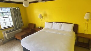Quarto de hotel com uma cama grande, um ventilador de teto, uma janela com cortinas, uma mesa com luminária e um aquecedor.