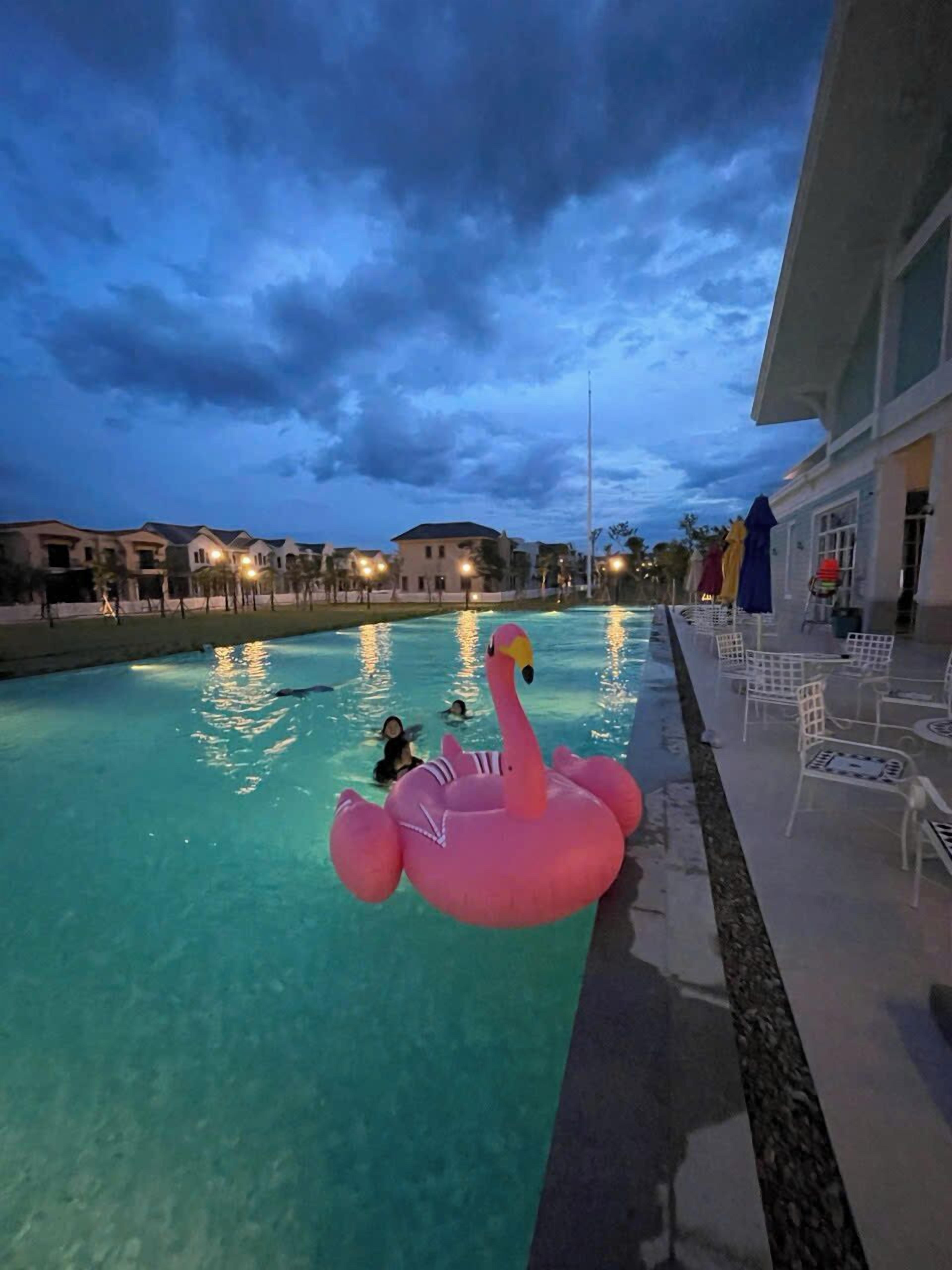 Piscina com um flutuador em formato de flamingo rosa, rodeada por espreguiçadeiras e um edifício com fachada branca.
