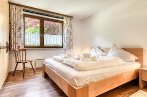 Quarto com cama de madeira, uma cadeira, janela com cortinas e vista para o verde lá fora.