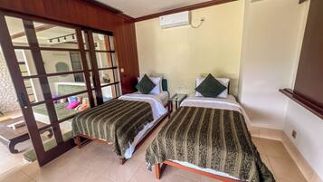 Quarto de hotel com duas camas, um separador de vidro, uma mesinha e um ar condicionado fixado na parede.