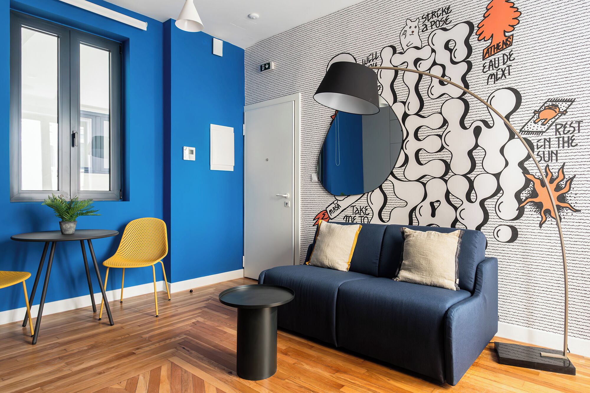 Uma sala de estar moderna com parede azul, um sofá com almofadas, uma mesa redonda, uma cadeira amarela e um abajur.
