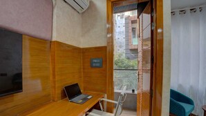 Um quarto pequeno com uma escrivaninha de madeira, um laptop, uma cadeira, uma janela e vista para edifícios lá fora.