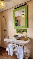 Uma casa de banho com parede revestida a azulejos verdes, uma pia branca com cuba, um chuveiro manual, um espelho e duas toalhas num suporte.