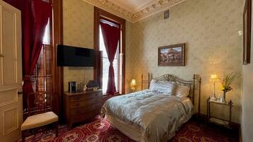 Quarto com cama, mesa de cabeceira, poltrona, cômoda, televisão e janela com cortinas.