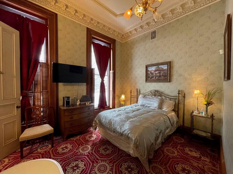Quarto com cama, mesa de cabeceira, poltrona, cômoda, televisão e janela com cortinas.