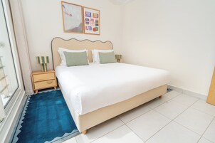 Uma cama bem arrumada com lençóis brancos, um tapete azul e uma mesa de cabeceira de madeira com abajur.