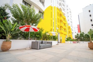 Uma área de estar ao ar livre com sofás listrados e guarda-sóis vermelhos e brancos, rodeada por vasos de plantas e um edifício amarelo ao fundo.