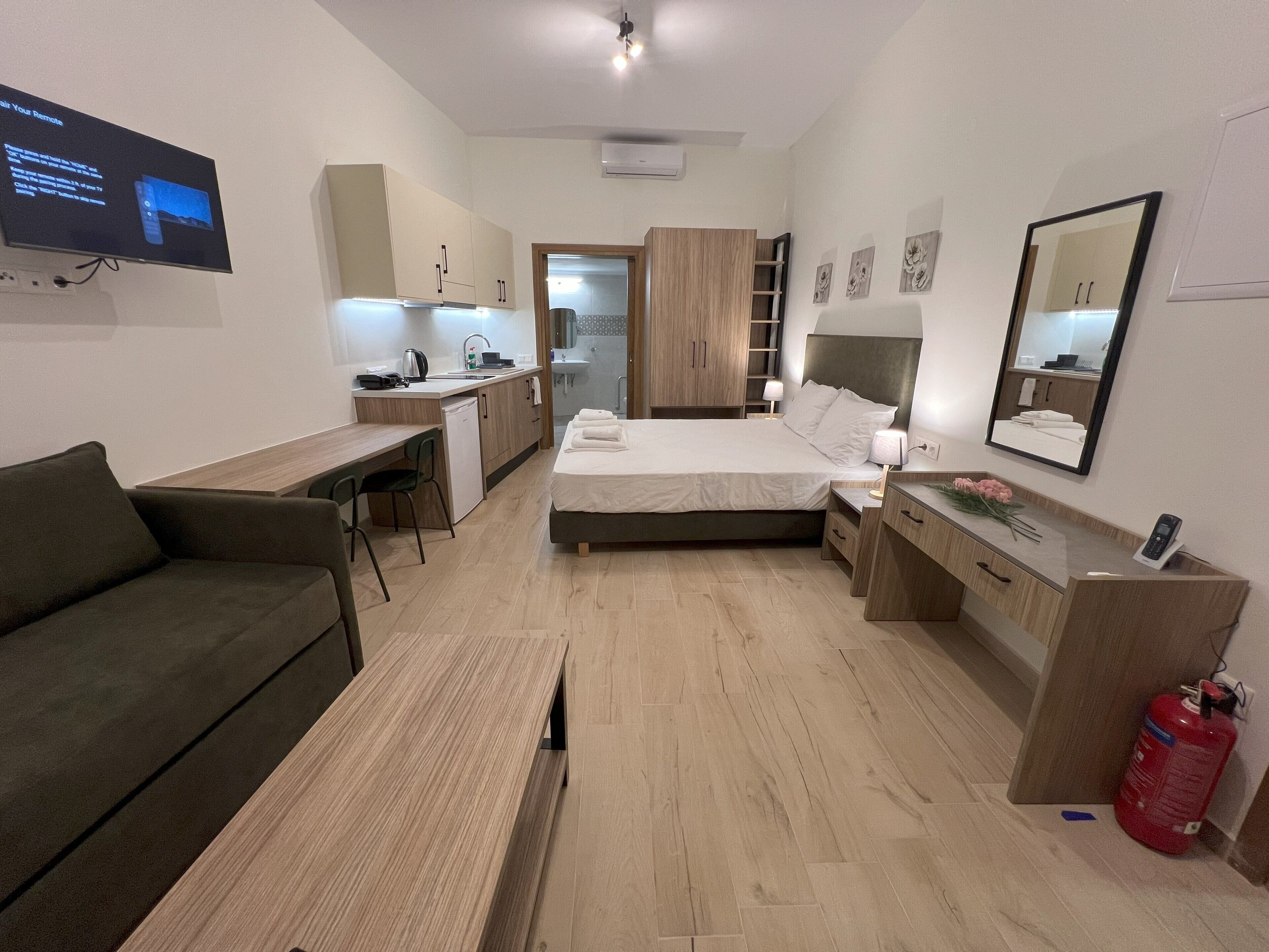 Quarto de hotel compacto com cama, escrivaninha, sofá, cozinha compacta e banheiro.