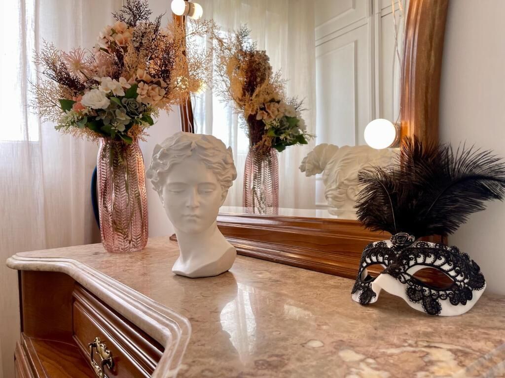 Uma bancada de mármore com uma escultura de busto, dois vasos com arranjos florais e uma máscara decorativa.
