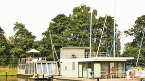 Uma casa flutuante atracada num cais, com um deque coberto e uma cabine pequena.