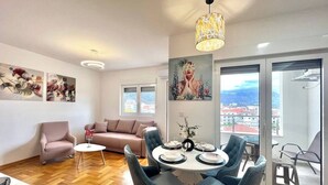 Uma sala de estar moderna com área de jantar, sofá, mesa de centro e lustre. Há duas janelas com vista para edifícios e montanhas.