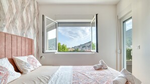 Quarto com cama grande, janela com vista aberta e paisagem urbana ao fundo.