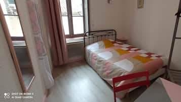 Quarto pequeno com uma cama de solteiro, piso de madeira, estrutura de cama de metal, uma cadeira vermelha, uma pequena mesa de cabeceira e janela com vista para o exterior.