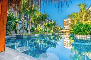 Uma piscina cristalina com espreguiçadeiras e guarda-sóis, cercada por vegetação exuberante e edifícios modernos.