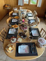 Mesa de madeira posta com diversos alimentos, incluindo pão, queijo e frutas, em um ambiente com paredes de madeira e uma janela.