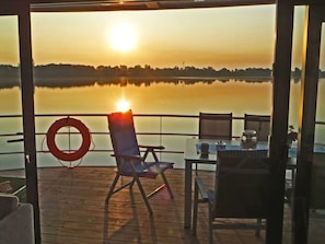 Um deck com mesa e cadeiras, com vista para um lago tranquilo ao pôr do sol.
