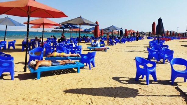 Uma praia com fileiras de cadeiras de plástico azuis e guarda-sóis vermelhos.