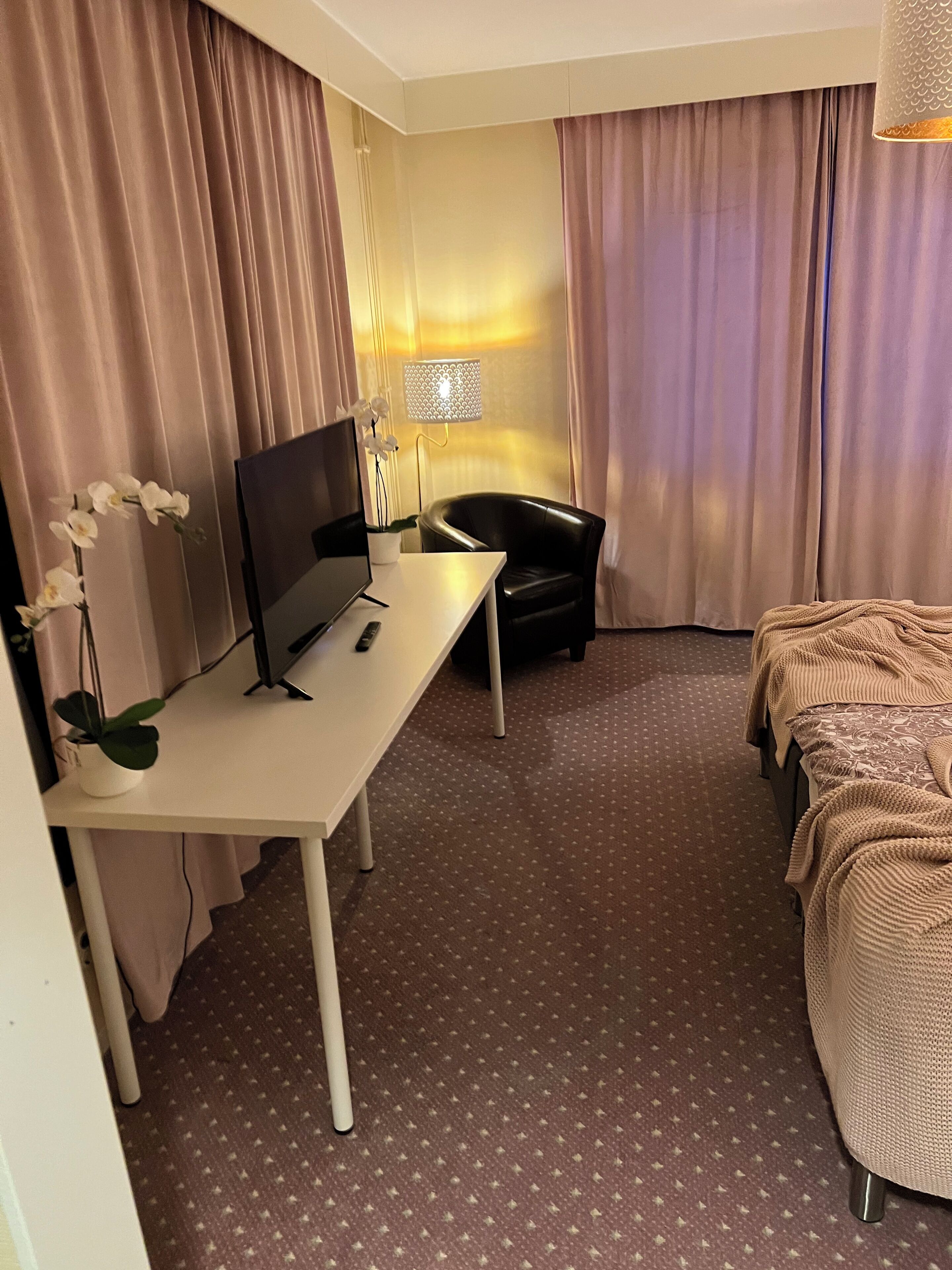 Quarto de hotel com uma cama, uma escrivaninha com computador, uma cadeira, um abajur e uma planta.