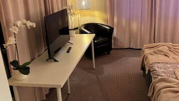 Quarto de hotel com uma cama, uma escrivaninha com computador, uma cadeira, um abajur e uma planta.
