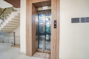 Um elevador moderno com estrutura de madeira e portas de vidro, situado num edifício com escadaria e guarda-corpo de vidro.
