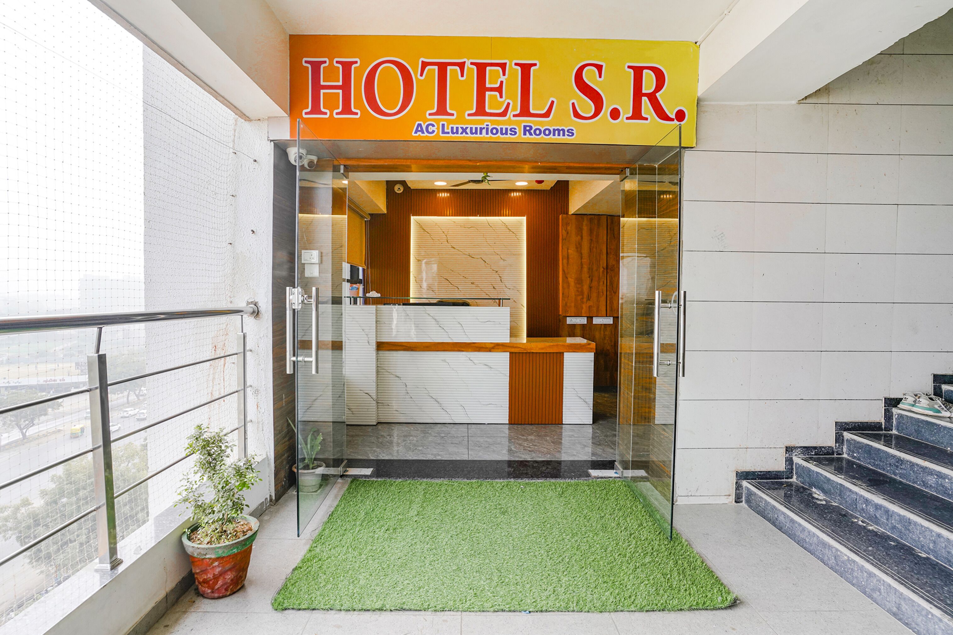 A entrada do HOTEL S.R. com um balcão de recepção, um tapete de grama artificial verde e um vaso de planta.