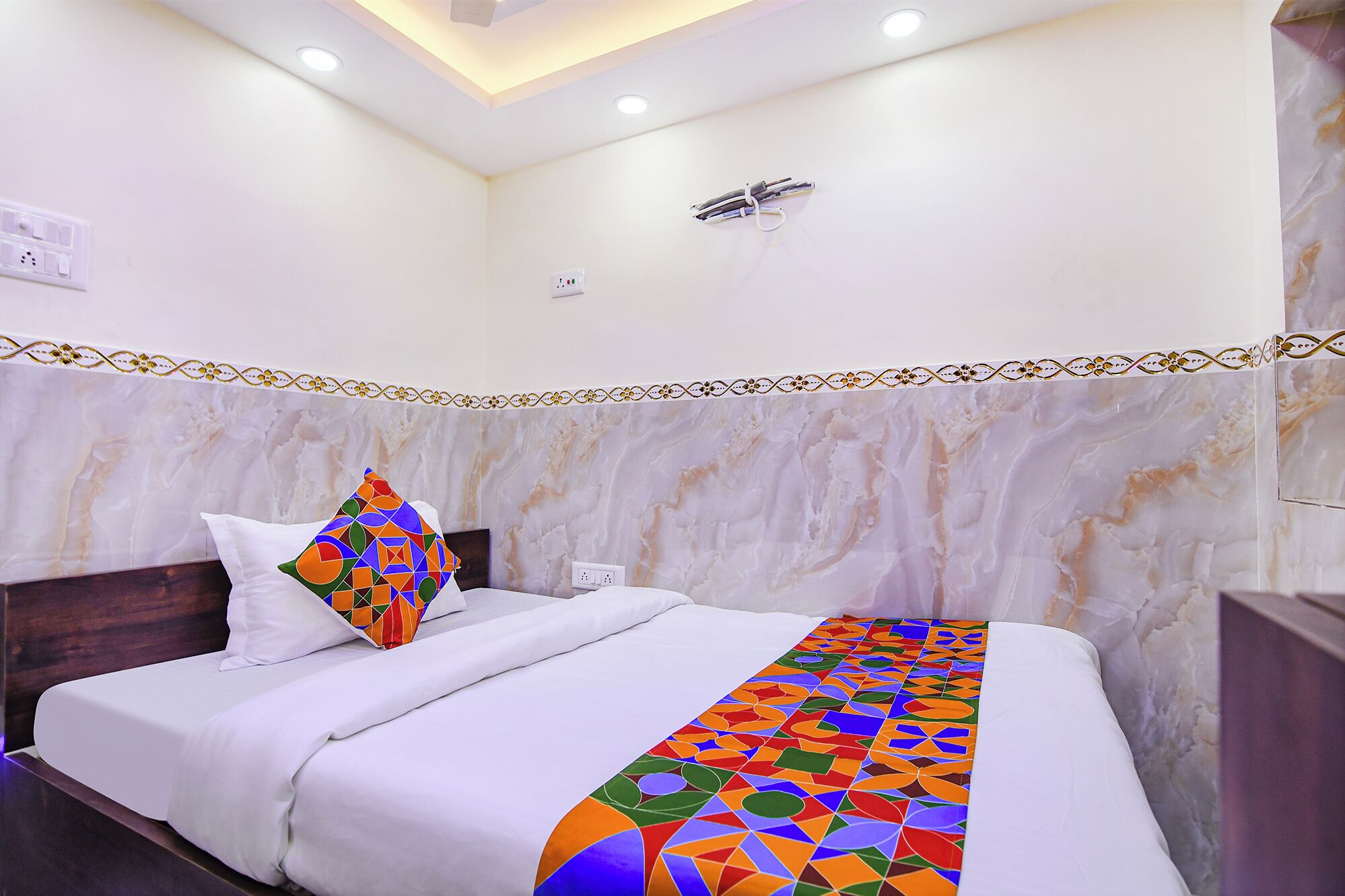 Quarto de hotel com cama, cabeceira, moldura decorativa e iluminação no teto.