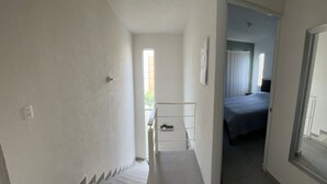 Um corredor que leva a um quarto com cama, janela com persianas e uma porta.