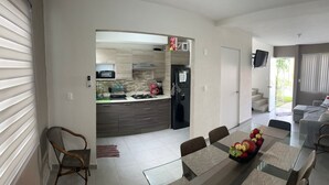 Cozinha moderna com armários escuros, micro-ondas e geladeira. Há uma área de refeições com mesa de vidro e cadeiras, e uma sala de estar com sofá e televisão.