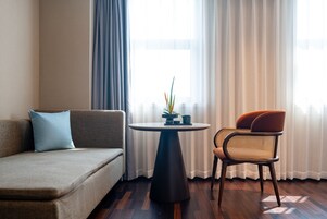 Um quarto com sofá, uma mesa redonda com uma planta, uma cadeira e uma janela com cortinas.