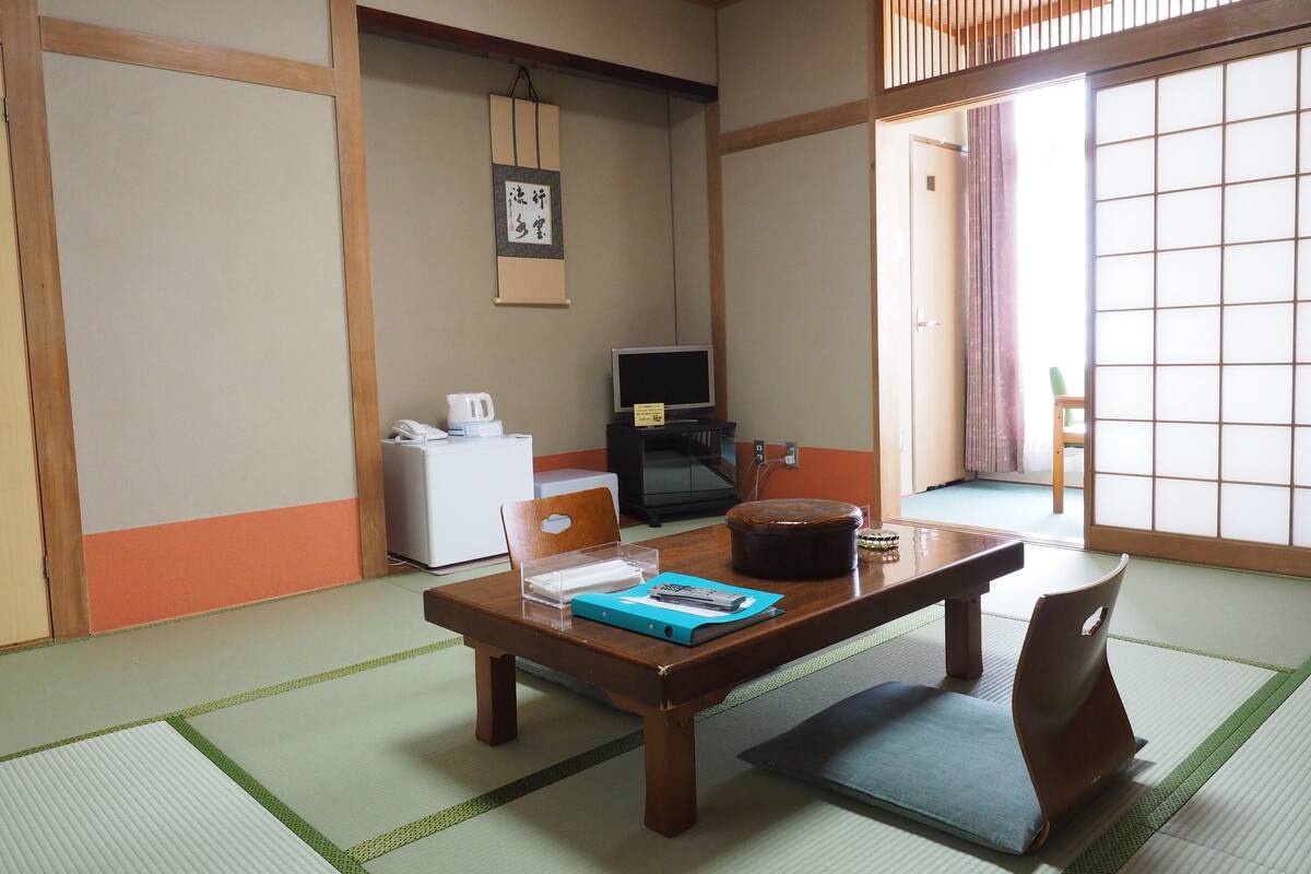Um quarto japonês tradicional com tatames, uma mesa de madeira com uma pasta azul, uma televisão e uma porta deslizante que dá para outro cômodo.