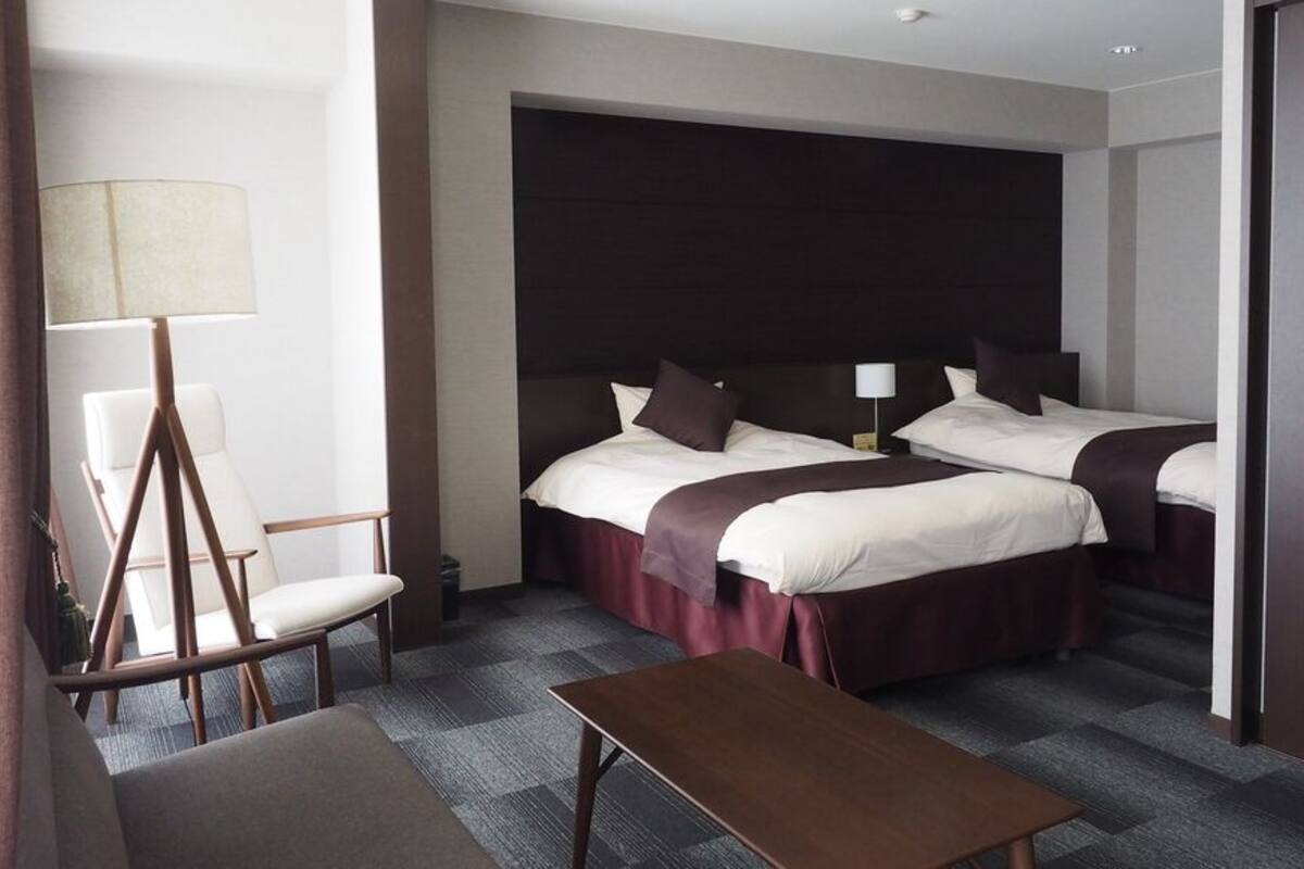 Quarto de hotel com duas camas, uma escrivaninha, uma cadeira e um abajur.