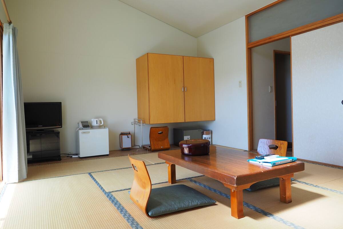 Um quarto japonês tradicional com tatames, uma mesa baixa, um armário de madeira, uma televisão, um frigorífico e uma pequena cozinha.