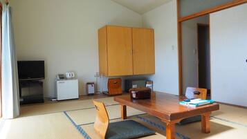 Um quarto japonês tradicional com tatames, uma mesa baixa, um armário de madeira, uma televisão, um frigorífico e uma pequena cozinha.