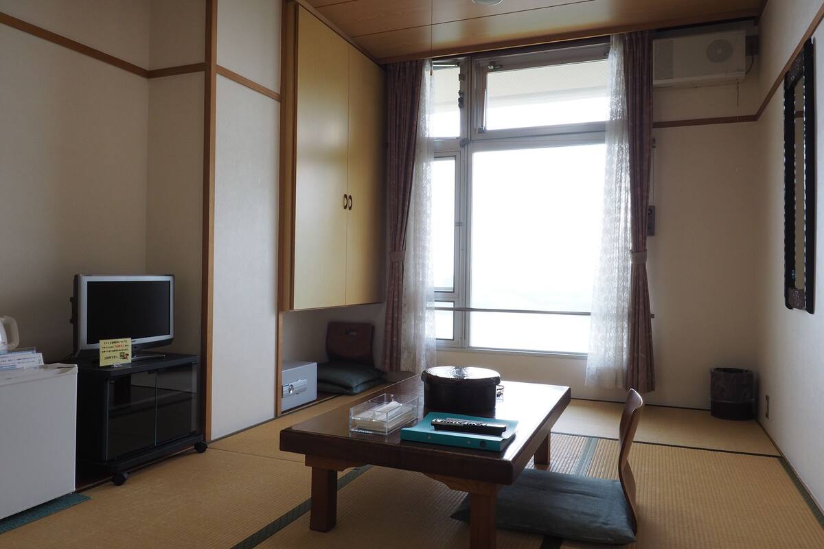 Um quarto japonês tradicional com tatames, uma mesa baixa com almofada, uma televisão sobre um suporte e uma porta deslizante com cortinas.