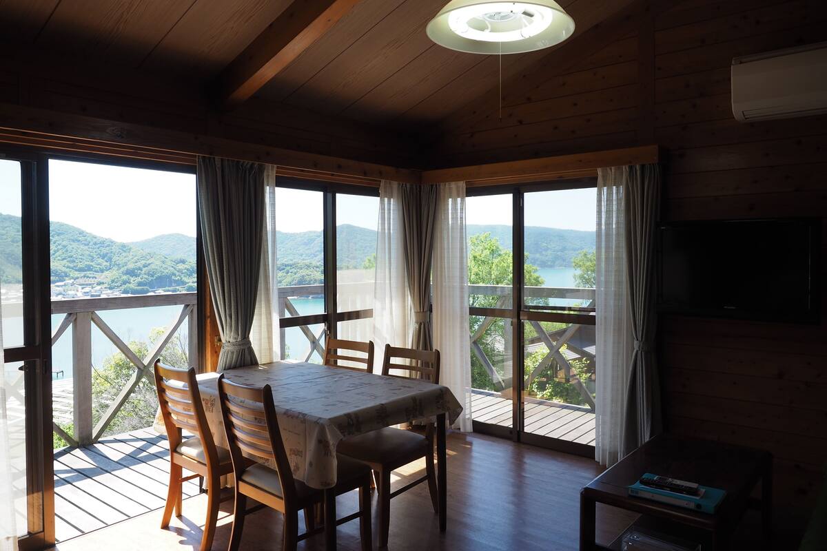 Interior em madeira com mesa e cadeiras, grandes janelas com cortinas e vista para um lago e montanhas.