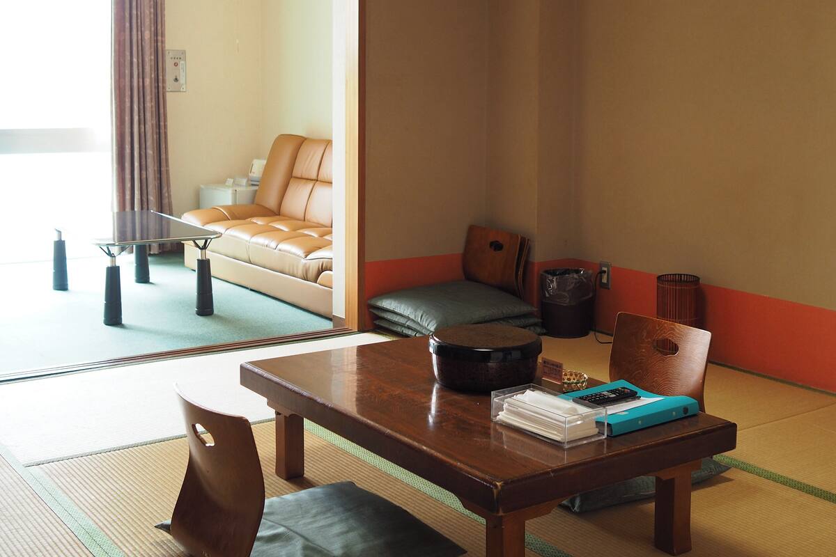 Um quarto japonês tradicional com tatames, uma mesa baixa com almofada, um sofá e uma cadeira de madeira.