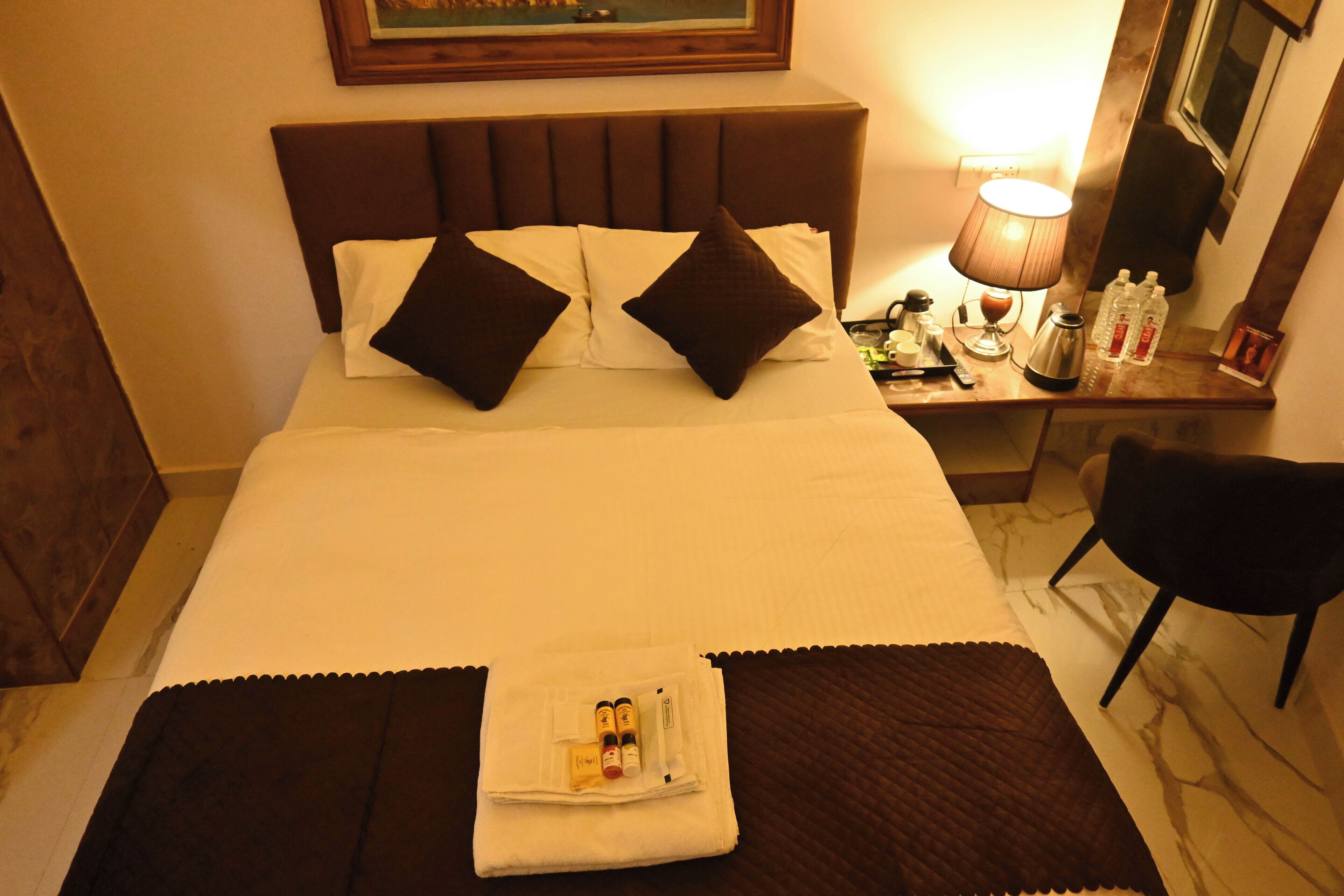 Quarto de hotel com uma cama grande, duas almofadas, mesa de cabeceira com abajur, uma cadeira e uma toalha dobrada com produtos de higiene pessoal sobre a cama.