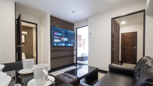 Quarto de hotel moderno com televisão de tela plana na parede, sofá de couro, mesa de jantar com pratos e talheres brancos, e vista para o quarto adjacente.