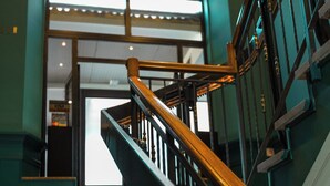 Uma escadaria com corrimão de madeira e balaústres de metal, conduzindo a um espaço interior iluminado com paredes verde-água e uma grande janela.