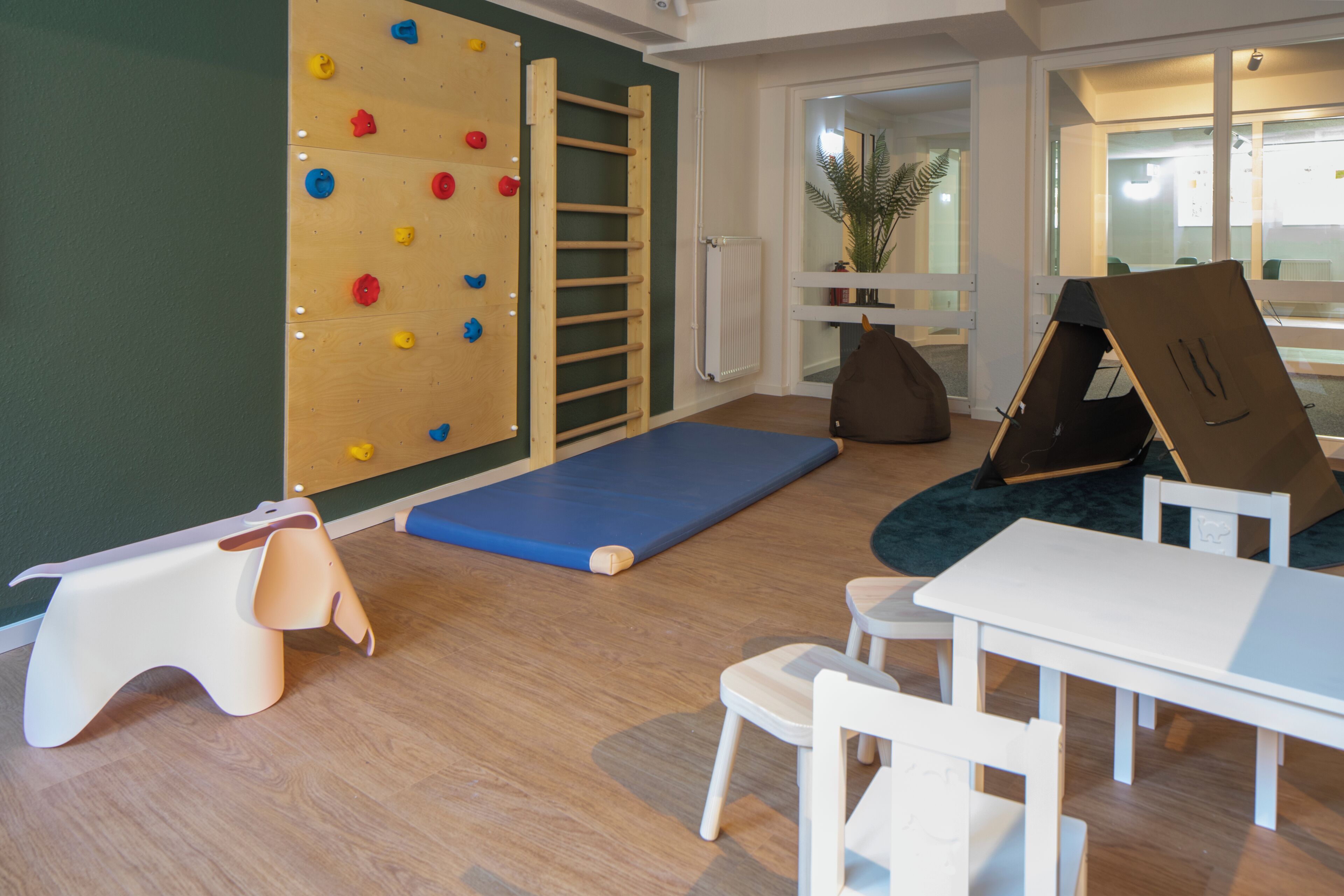 Uma sala de brincadeiras com parede de escalada, tapete macio para as crianças e uma mesa pequena com cadeiras.