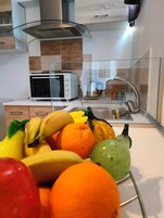 Uma cozinha com micro-ondas, pia e uma fruteira com bananas, laranjas e uma fruta verde.