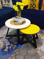 Uma mesa redonda branca com um banquinho amarelo, um vaso com flores amarelas e um livro intitulado 'CHANEL' sobre a mesa.